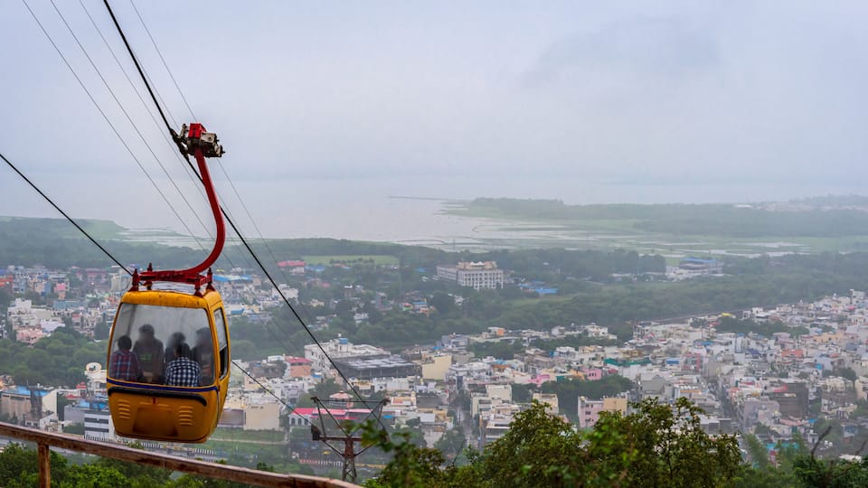 Um teleférico com passageiros, paisagem urbana e um rio ao fundo.
