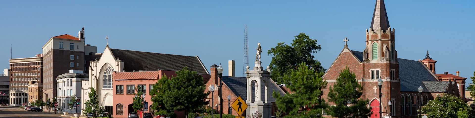 Uma praça urbana com uma igreja imponente, uma estátua e edifícios ao redor.