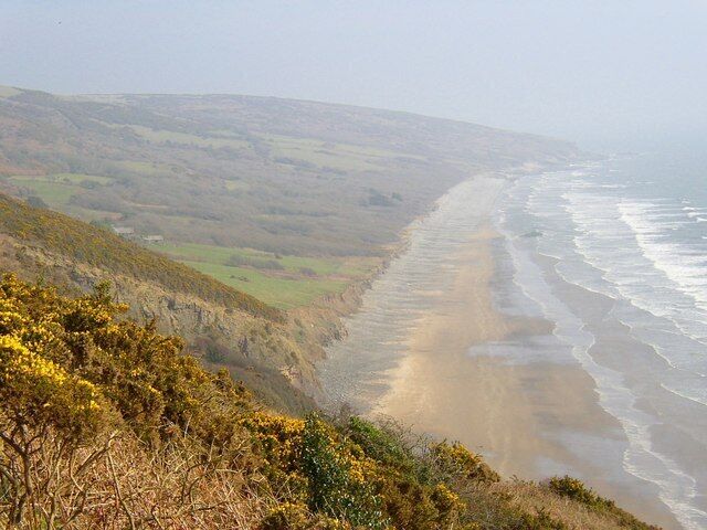 Paisagem costeira com praia arenosa, colinas ondulantes e vegetação escassa.