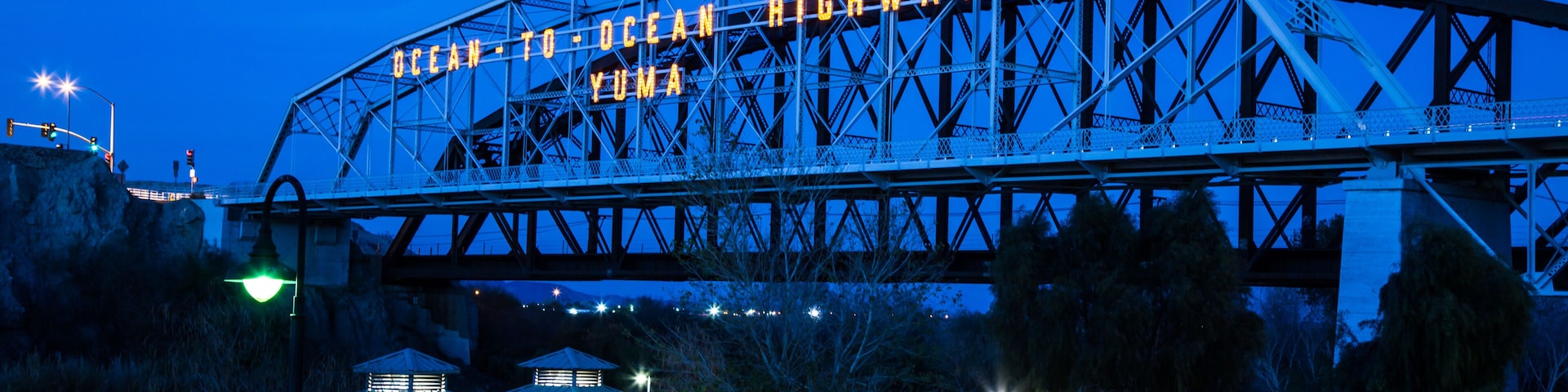 Uma ponte de aço com placas iluminadas que dizem 'OCEAN TO OCEAN HIGHWAY YUMA', rodeada por um parque com árvores e um coreto.