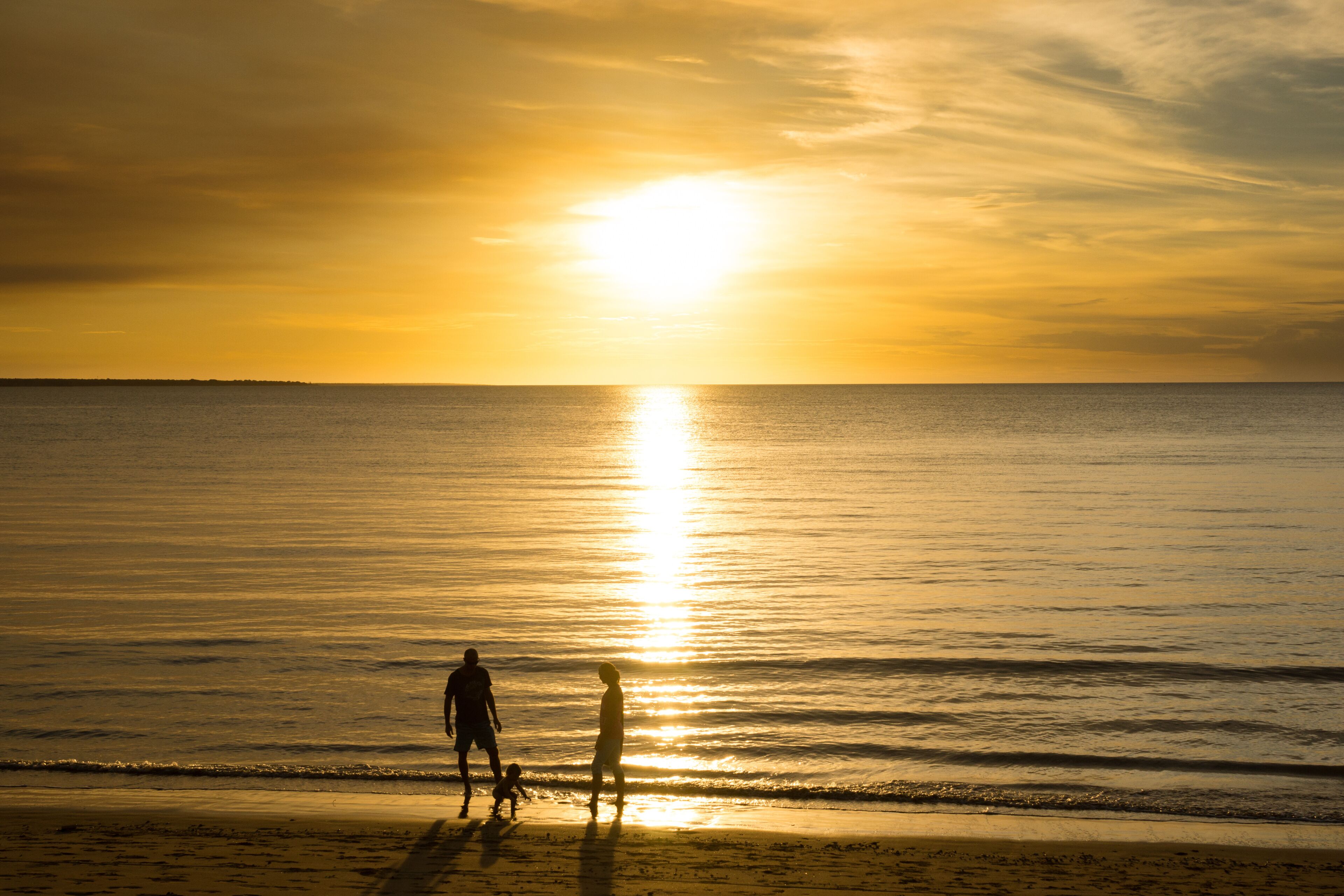 Um pôr do sol sobre um mar calmo, com silhuetas de pessoas e uma criança na praia.