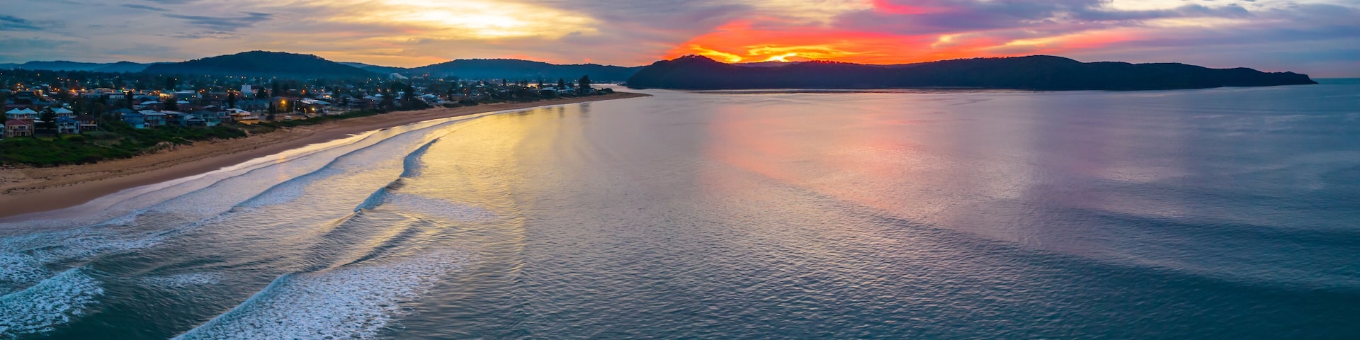Paisagem costeira com um pôr do sol, mar calmo e uma cidade à beira-mar.