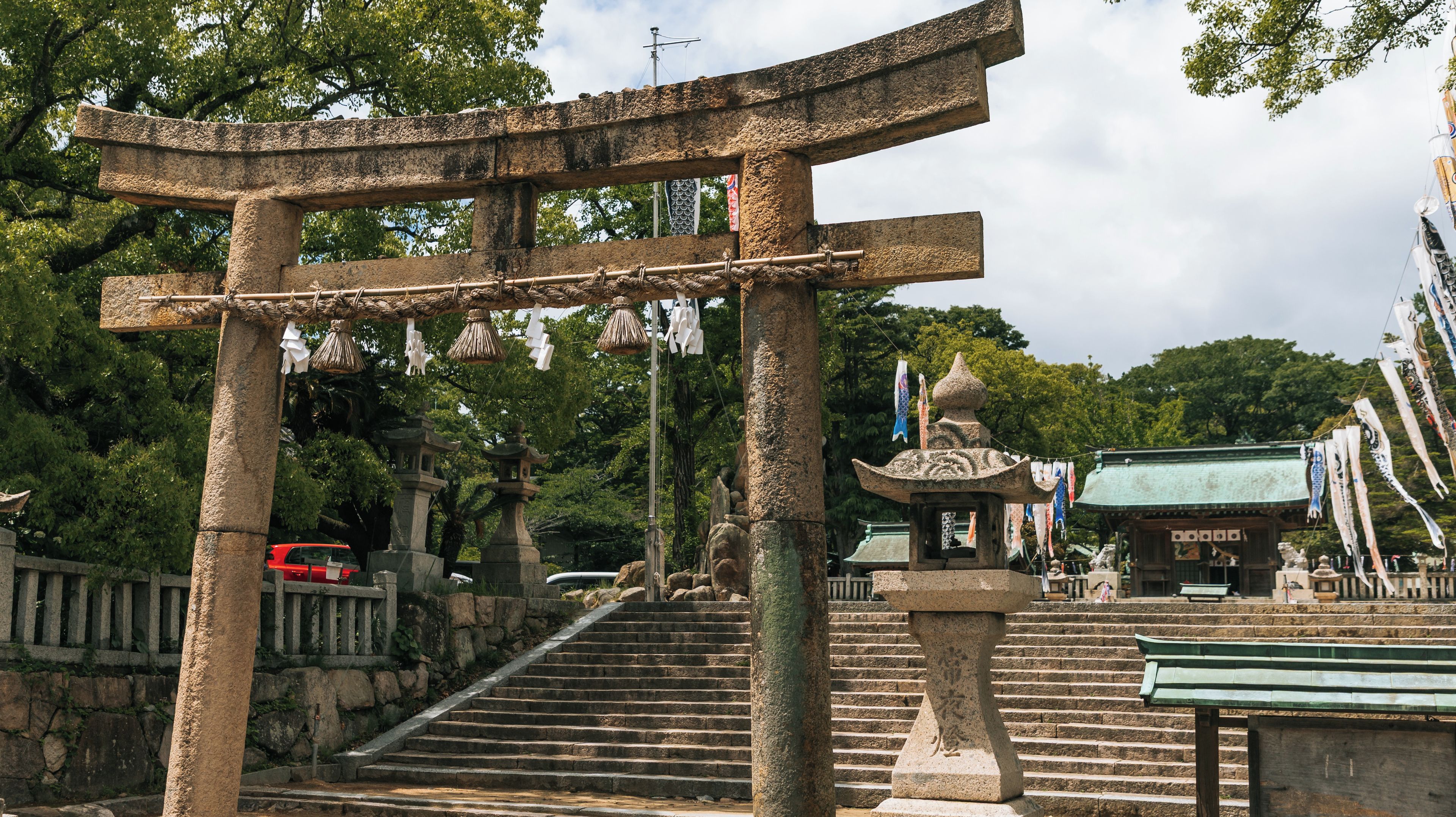 Um portão torii de pedra que dá acesso a um santuário, com uma pequena pagoda e um edifício tradicional ao fundo.