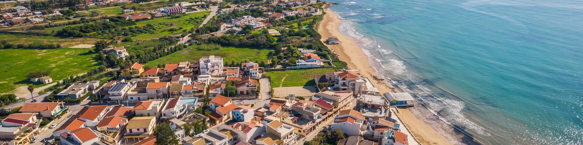 Vista aérea de uma vila costeira com praia, edifícios residenciais e campos verdes.