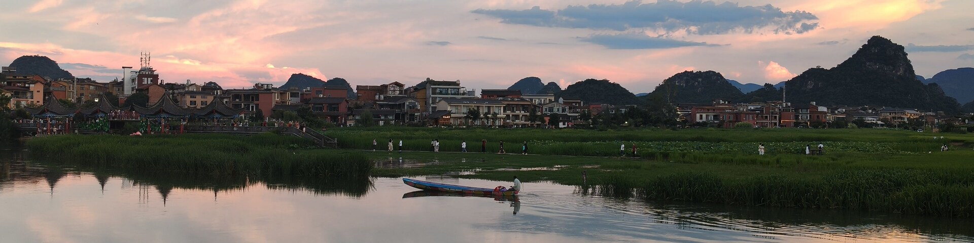 Um rio calmo refletindo o céu, um barco tradicional e uma cidade com edifícios e montanhas ao fundo.