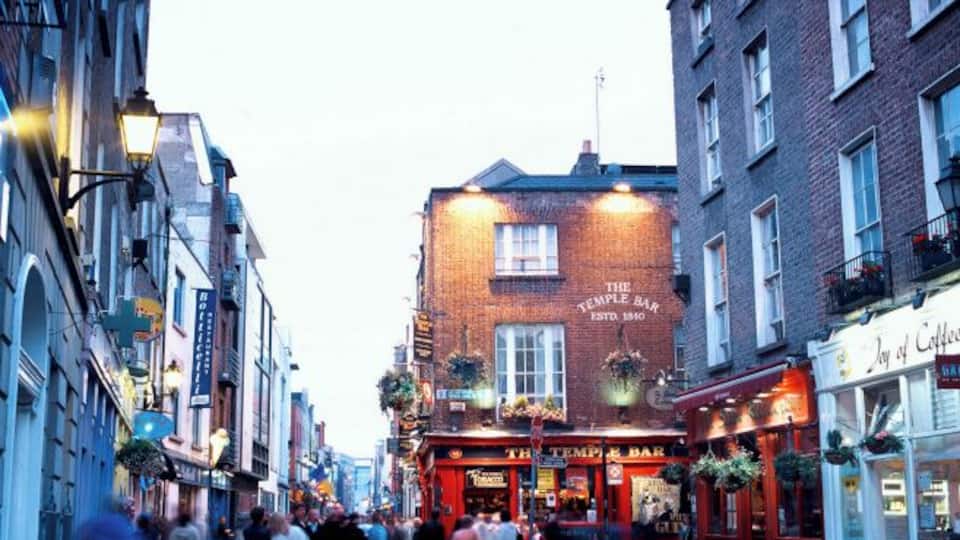 Rua de paralelepípedos com edifícios históricos, incluindo The Temple Bar e uma cafeteria.