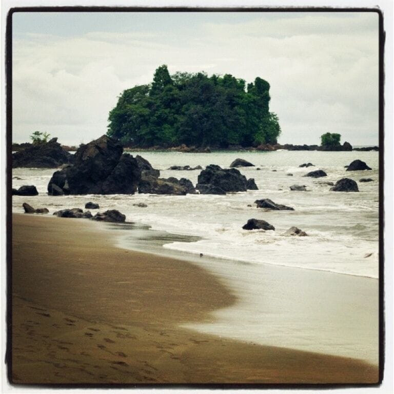 Uma praia com pedras grandes e uma pequena ilha com vegetação densa ao fundo.