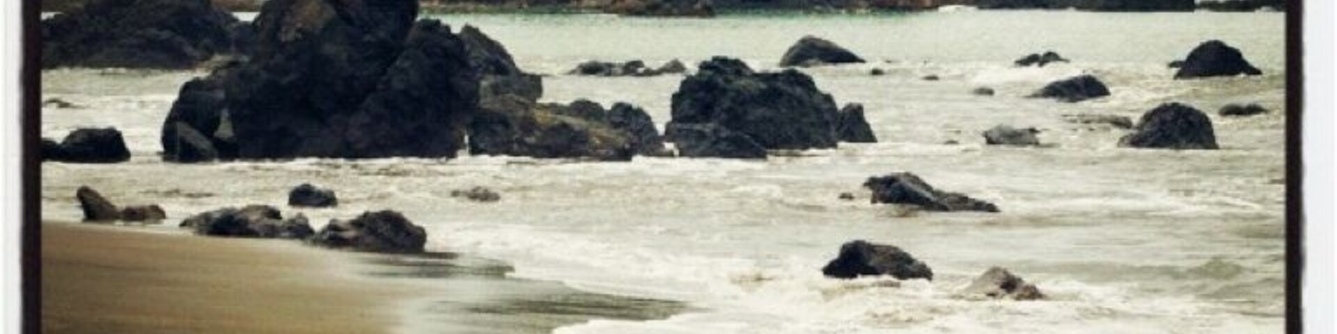 Uma praia com pedras grandes e uma pequena ilha com vegetação densa ao fundo.