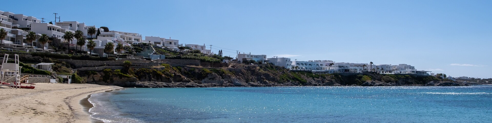 Uma praia com águas azul-claras, areal arenoso e edifícios brancos numa encosta.