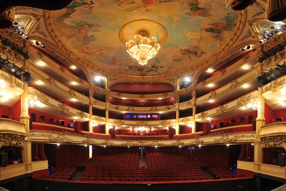 Um teatro ornamentado com assentos vermelhos, um candelabro e um mural no teto.