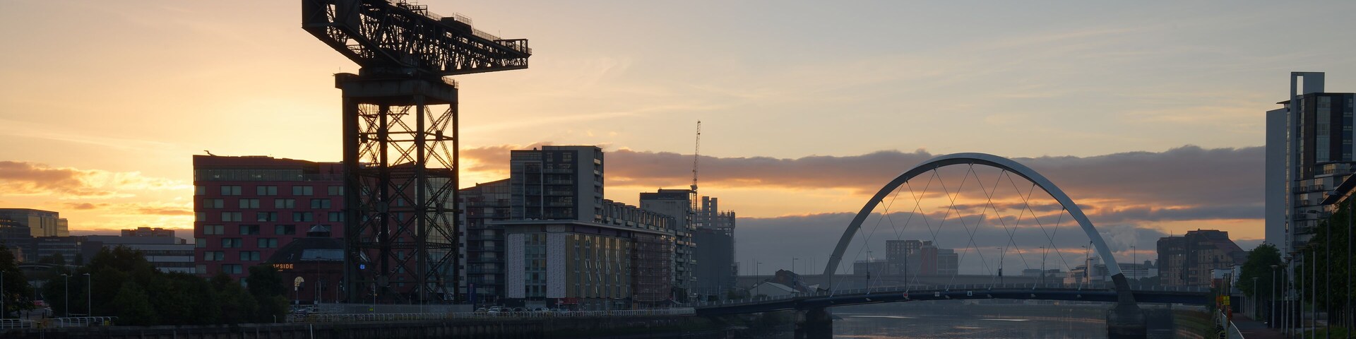 Um pôr do sol sobre um rio, com uma ponte e edifícios industriais.
