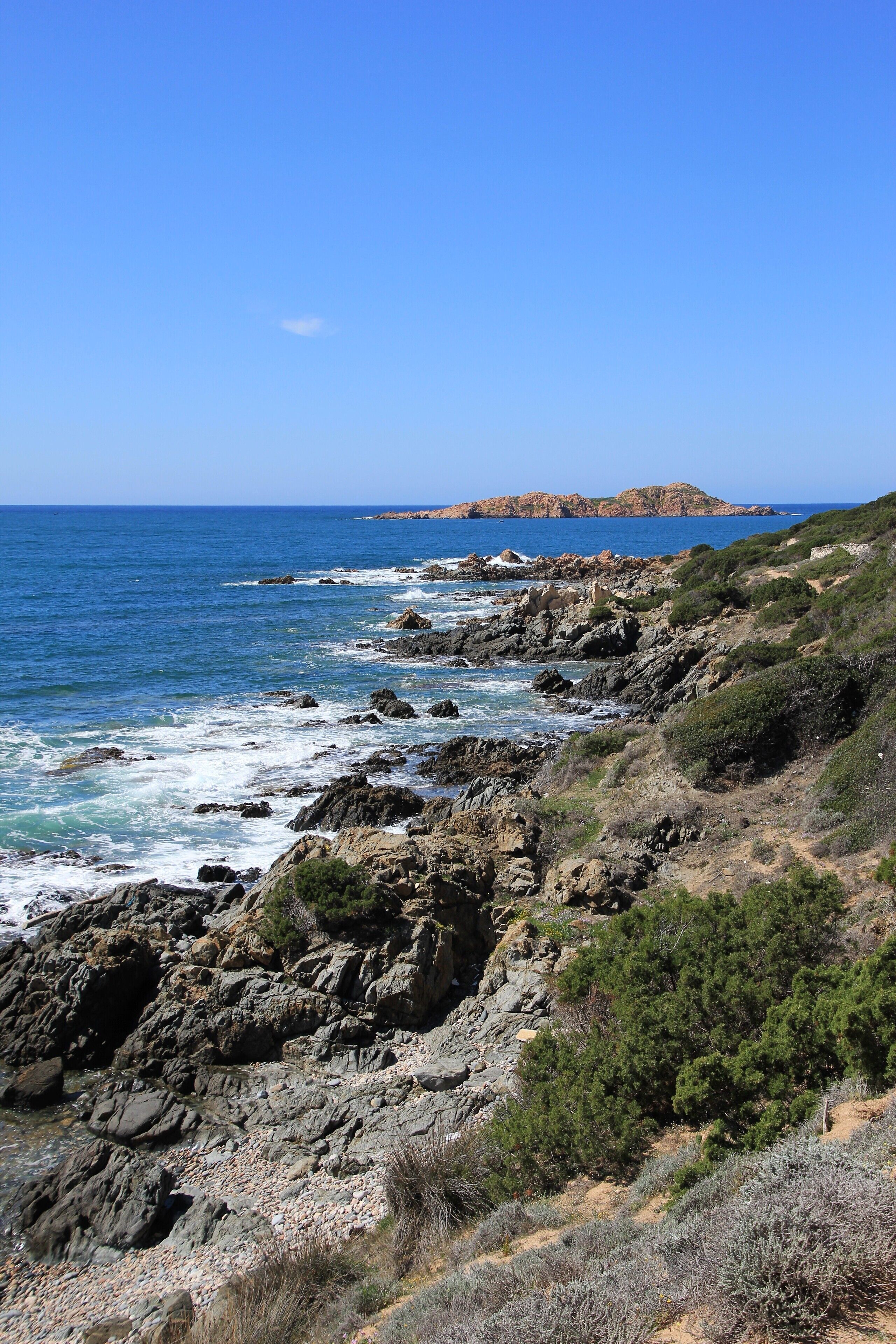 Litoral rochoso com ondas quebrando na costa, uma ilha distante e vegetação escassa.