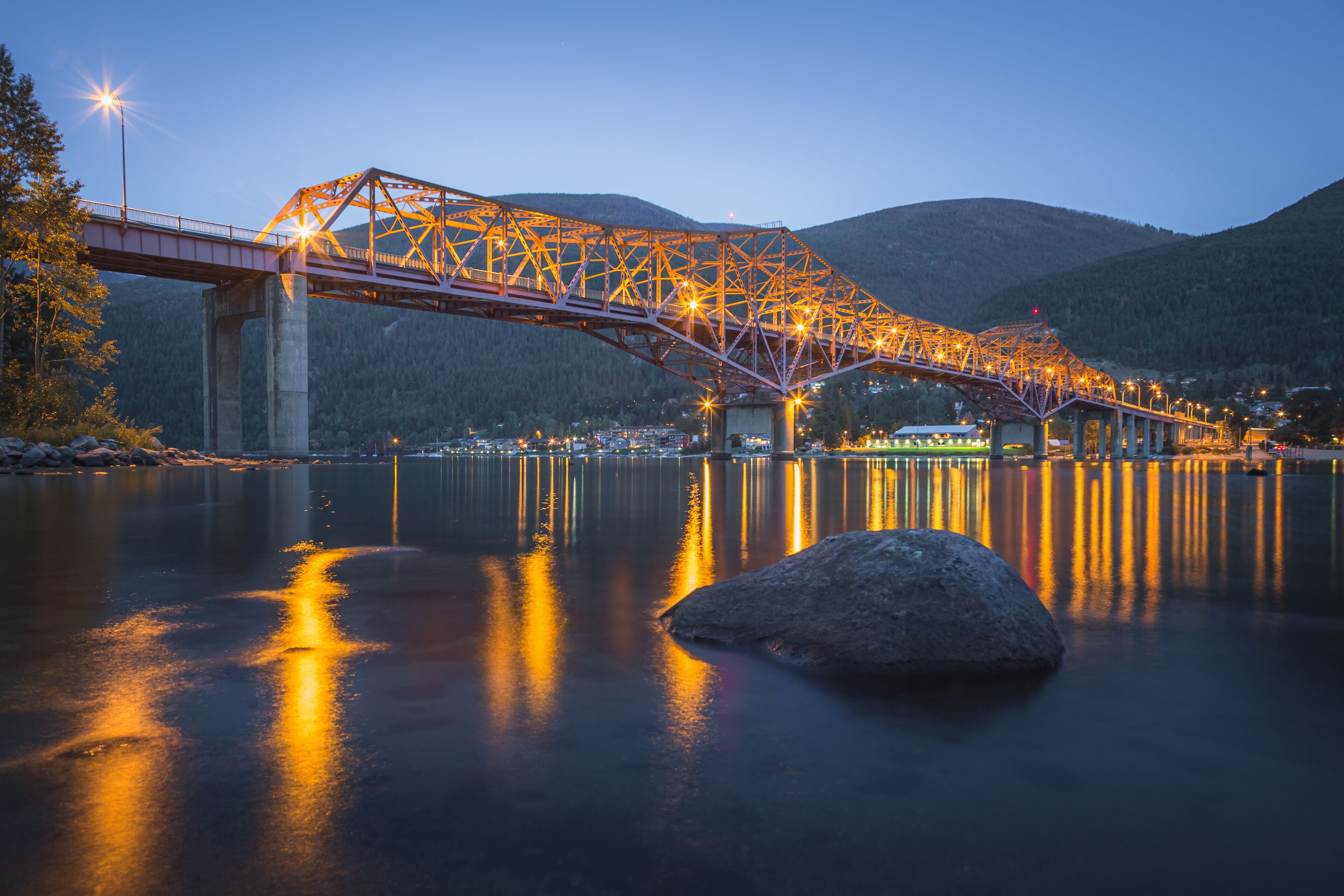Uma ponte de aço com luzes refletindo em um rio calmo, rodeada por montanhas.