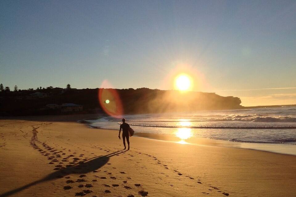Uma praia com pegadas indo em direção à água, uma pessoa com prancha de surf e o sol se pondo sobre o oceano.