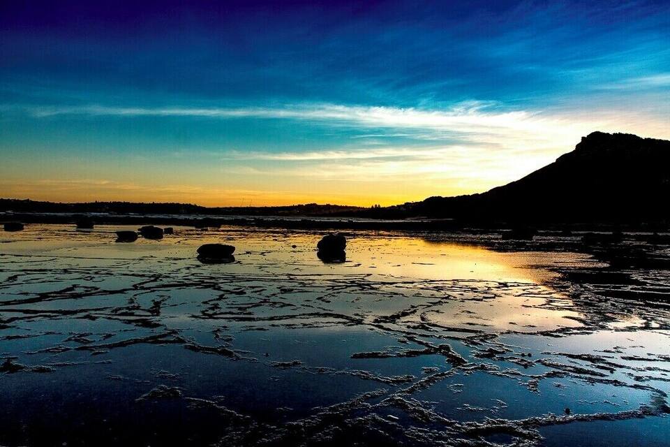 Pôr do sol sobre uma paisagem alagada com rochas e uma montanha ao fundo.