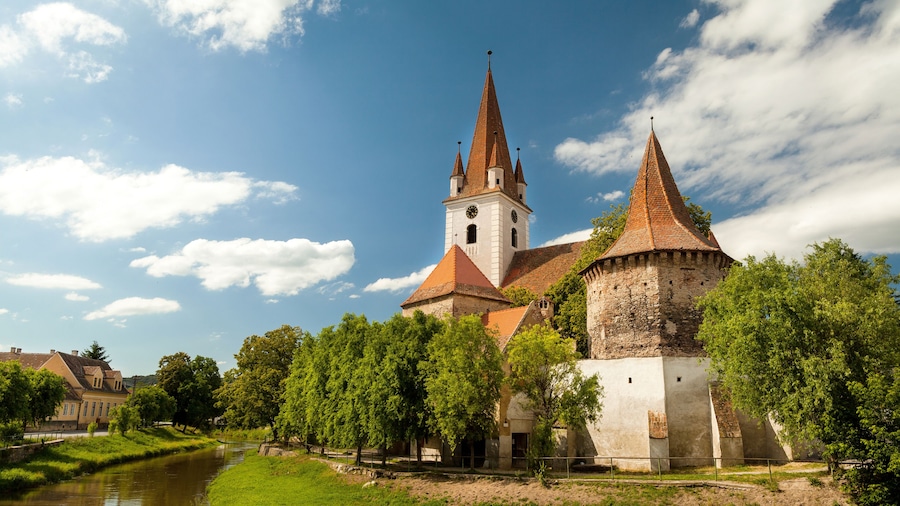 Uma igreja histórica com uma torre alta e uma torre menor adjacente, cercada por vegetação exuberante e um rio.