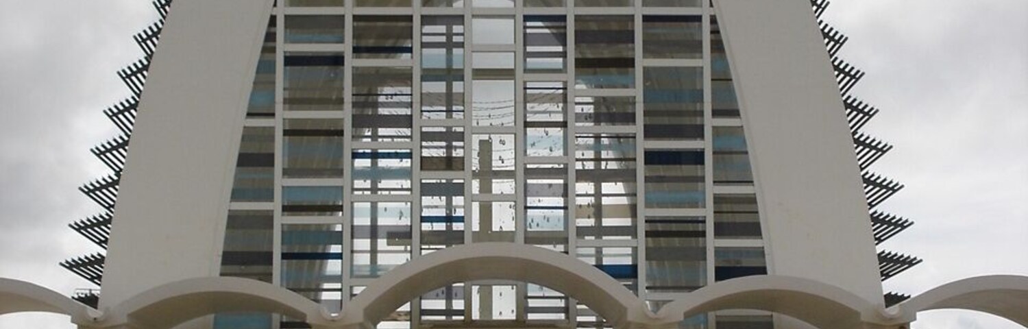 Um edifício moderno com uma grande entrada em arco, rodeado por vasos de plantas e um céu claro.