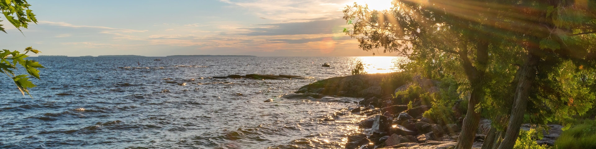 Uma costa rochosa com o sol refletindo na água, cercada por vegetação e árvores.