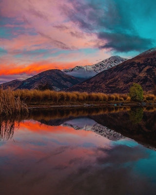 Uma cordilheira com picos nevados, um lago tranquilo refletindo o céu e um primeiro plano de gramíneas altas.