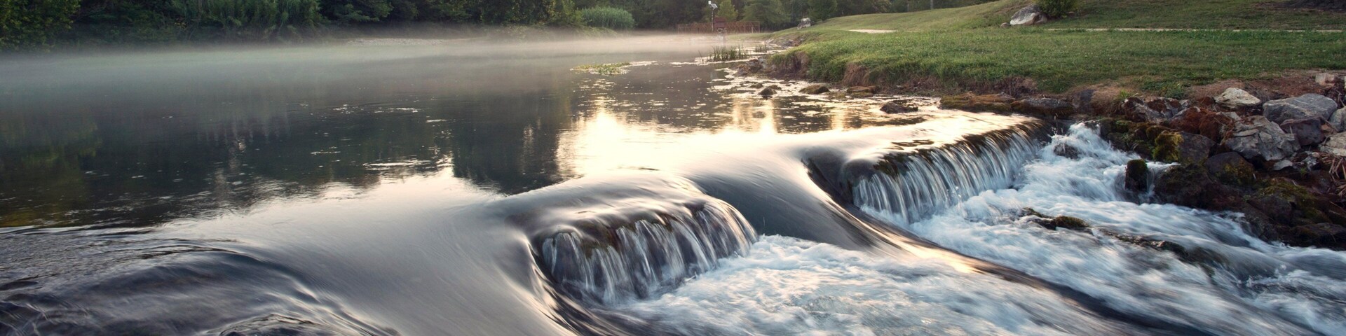 Uma cascata despencando sobre pedras em um rio calmo, cercada por árvores e uma área gramada.