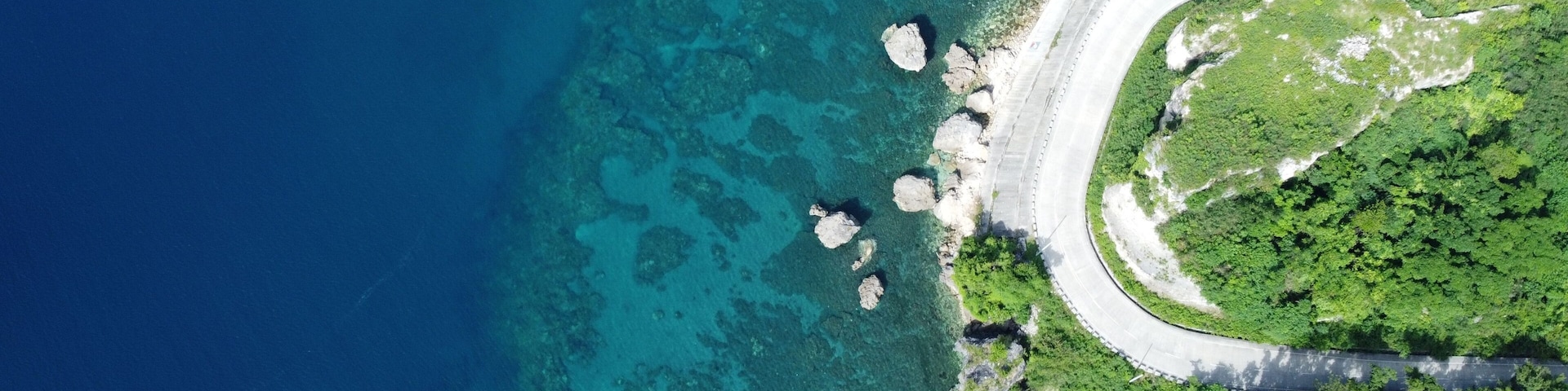 Uma estrada costeira serpenteia por uma orla rochosa, com águas azul-claras e vegetação exuberante.