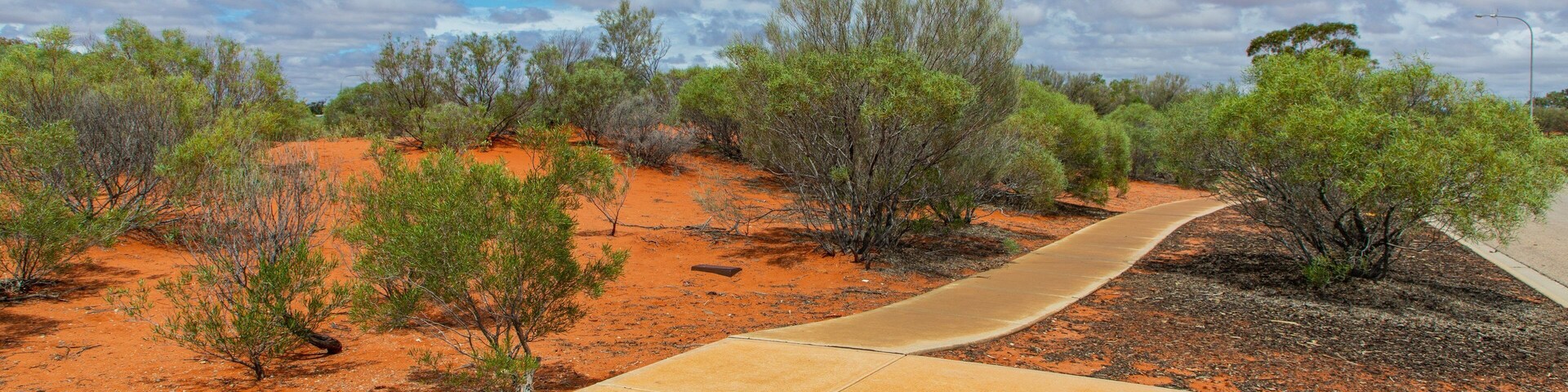 Um caminho de concreto sinuoso atravessa uma paisagem desértica avermelhada com vegetação verde escassa e céu parcialmente nublado.