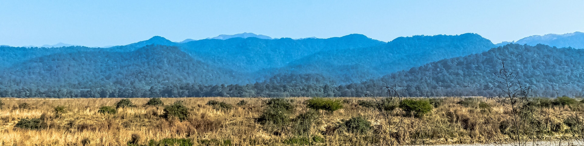 Um rio com leito pedregoso, cercado por pastagem seca e montanhas distantes sob um céu claro.