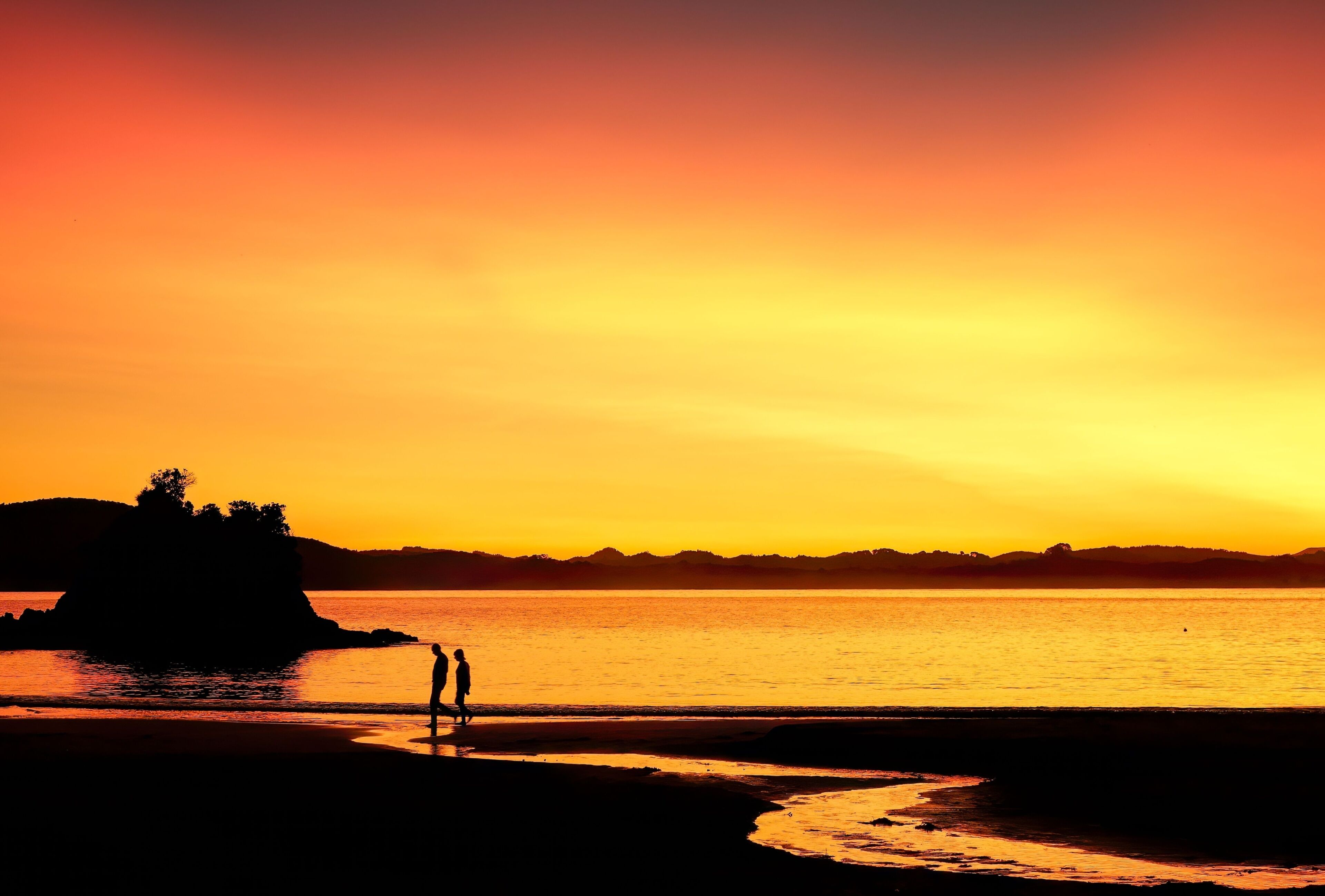 Um pôr do sol sobre uma praia com duas silhuetas de pessoas à beira-mar.
