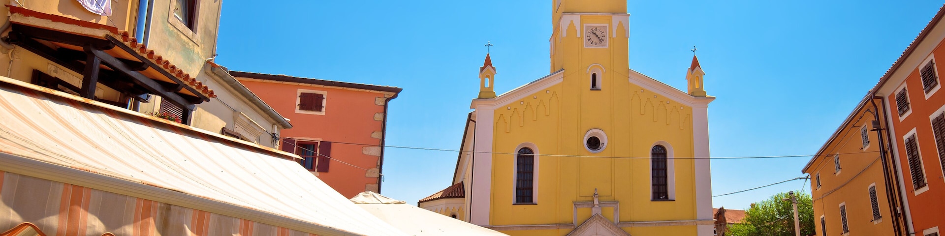 Uma igreja amarela com um campanário alto, toldo branco e carros estacionados na rua.