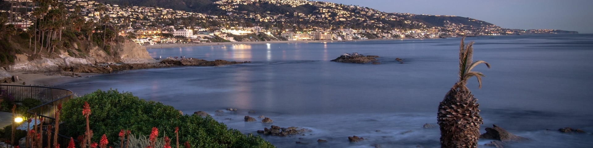 Paisagem costeira ao entardecer, com vista para o oceano e uma colina com edifícios iluminados.