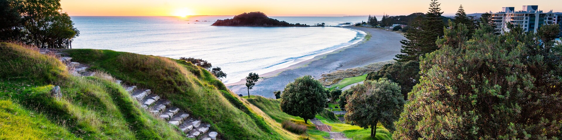 Paisagem costeira com um morro gramado, uma praia e um pôr do sol.