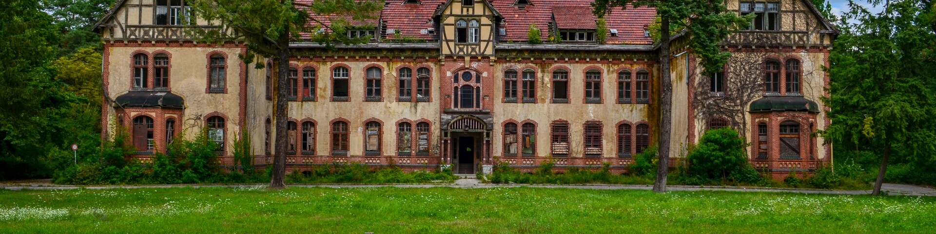 Um edifício grande e histórico com telhado de telhas vermelhas, rodeado por árvores verdes exuberantes e um gramado.
