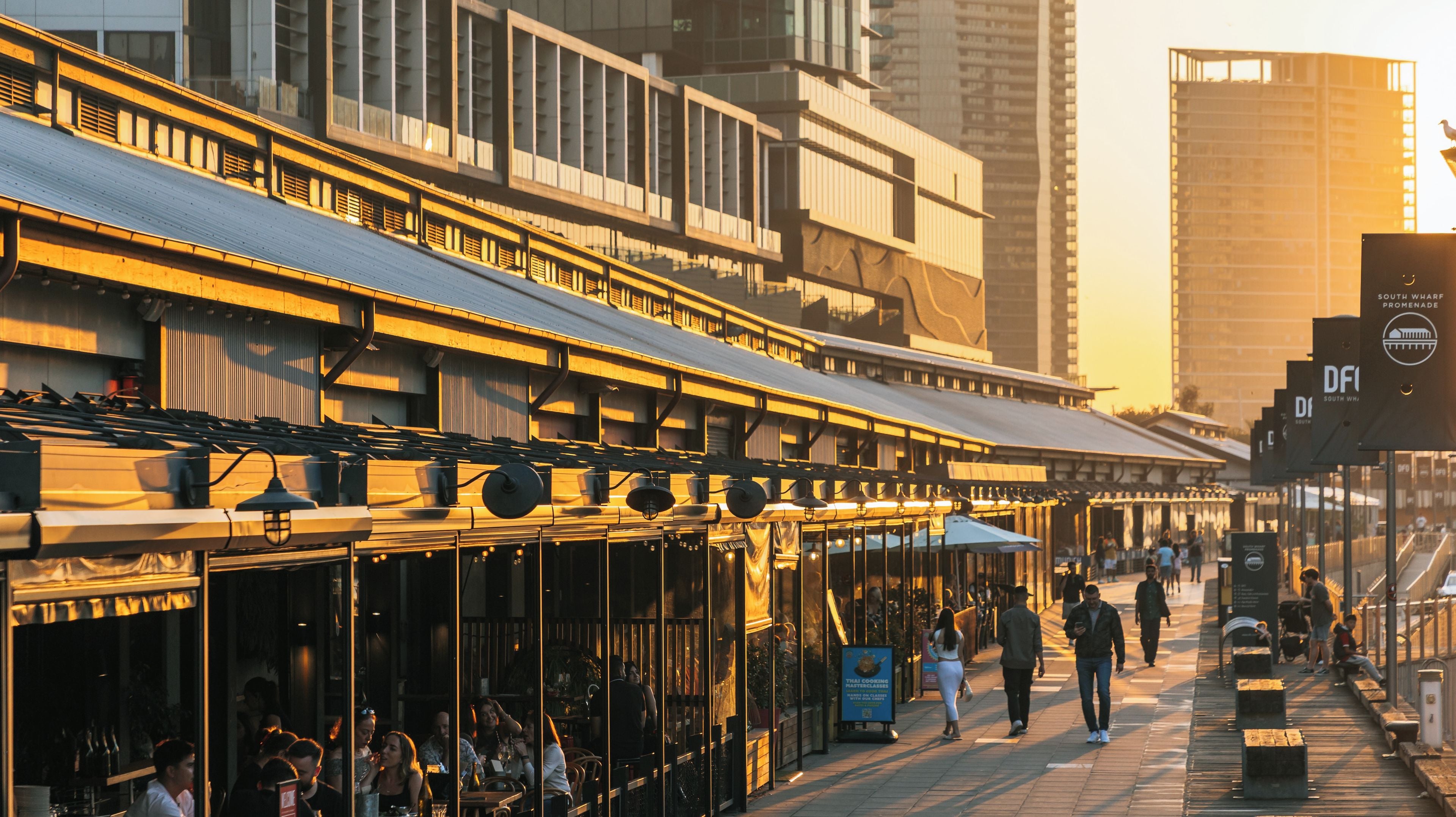Uma rua urbana movimentada com uma fileira de cafés ao ar livre, pedestres e edifícios modernos.