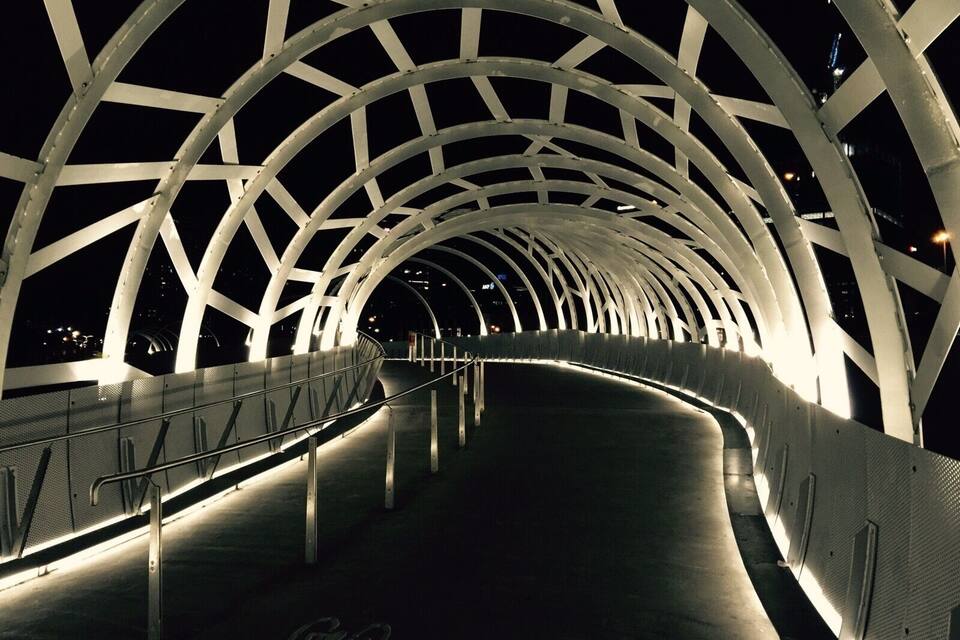 Vista noturna de uma ponte para pedestres curva, com vigas e grades brancas.