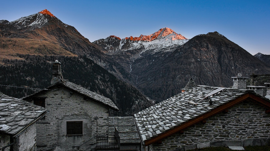 Uma aldeia de montanha com edifícios de pedra e um pico proeminente com manchas de neve laranja.