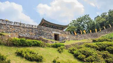 Uma fortaleza coreana tradicional com muros de pedra e telhado de telhas, rodeada por vegetação exuberante e árvores.