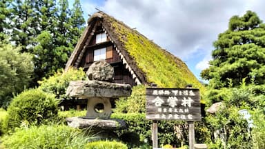 Uma casa tradicional japonesa com telhado de palha e um candeeiro de pedra à frente, rodeada por vegetação exuberante.