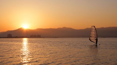 Pôr do sol sobre águas calmas, com o perfil de montanhas ao fundo e uma pessoa praticando windsurf.
