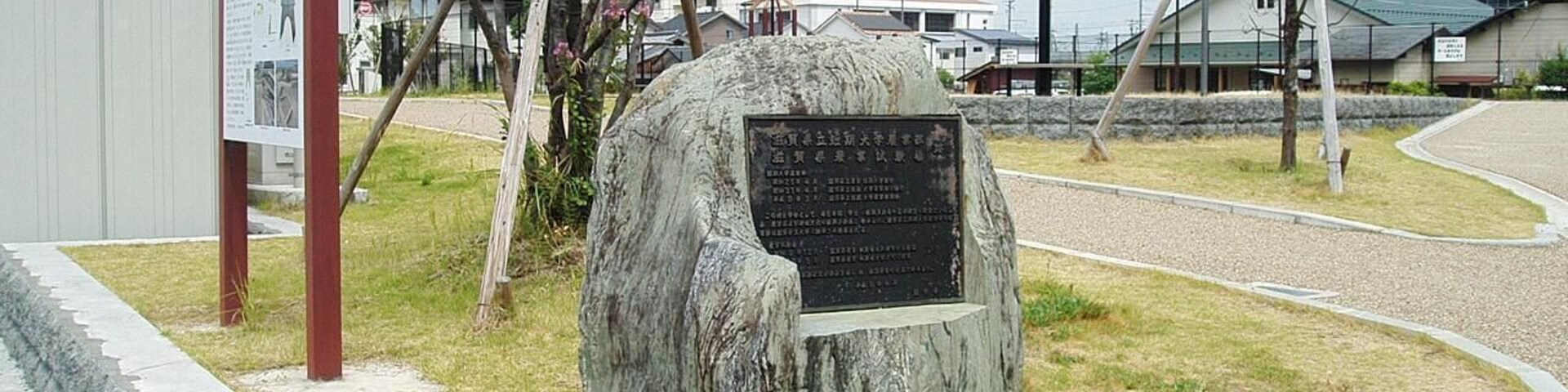 Um monumento de pedra com texto gravado, rodeado por um caminho de cascalho e relva escassa, com edifícios e árvores ao fundo.
