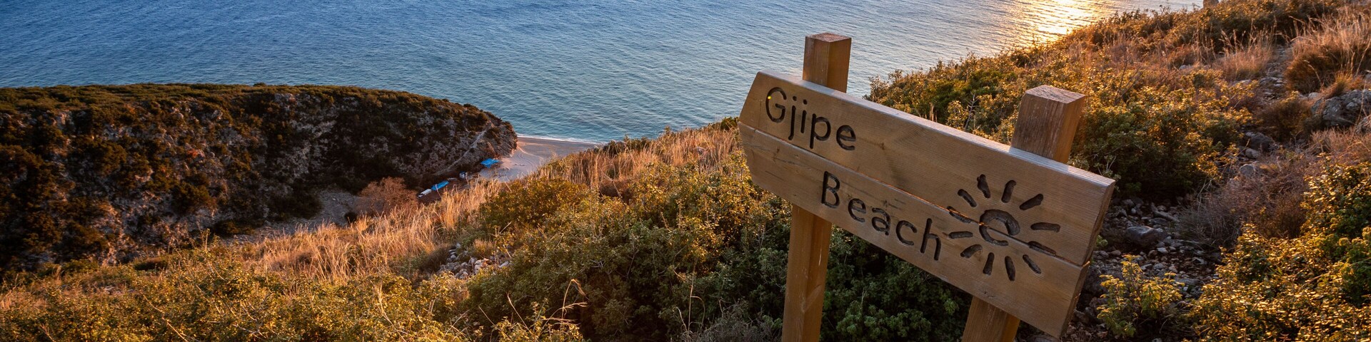 Um sinal de madeira com a inscrição 'Gijpe Beach', com vista para a paisagem costeira, o oceano e o pôr do sol.