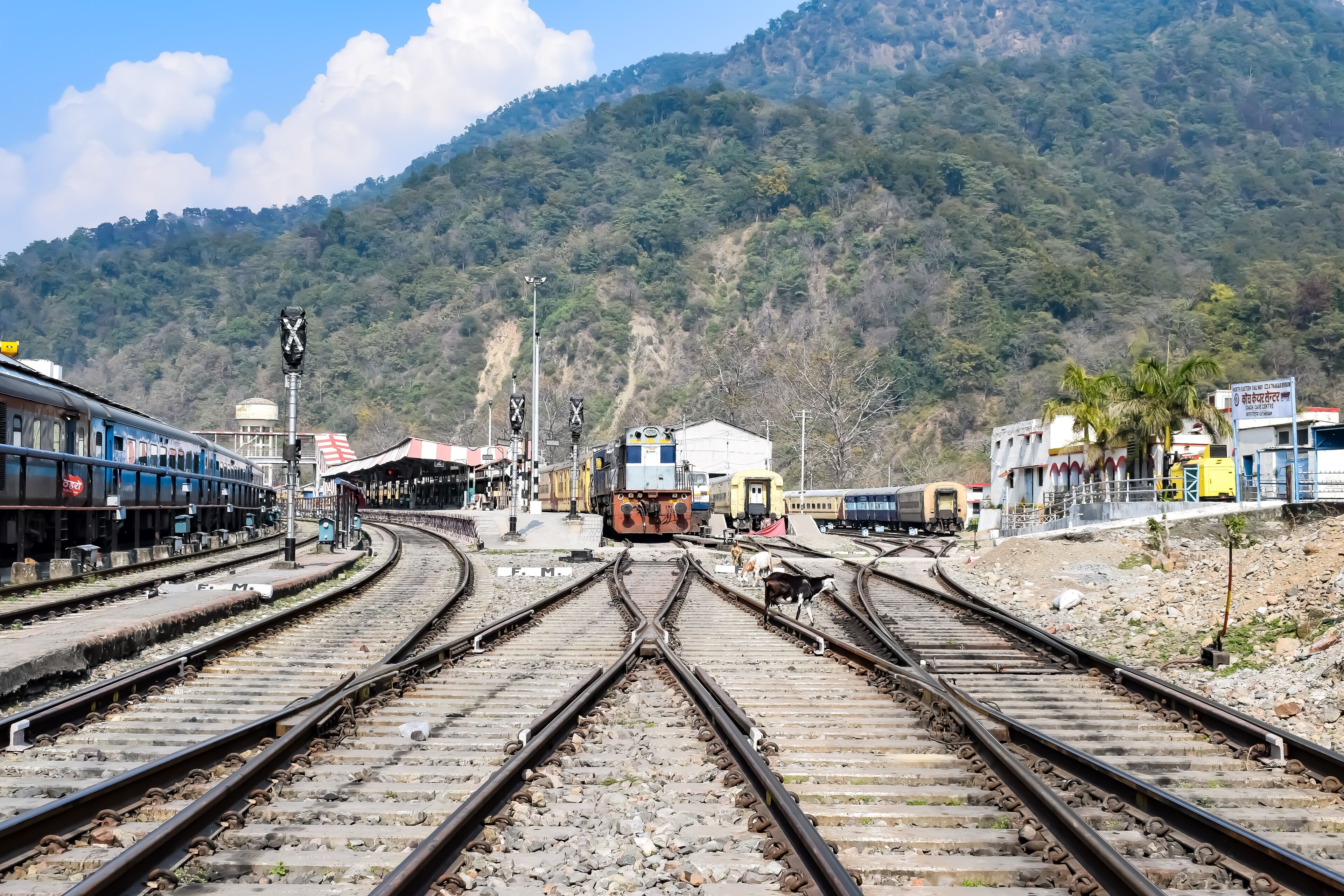 Um entroncamento ferroviário com várias linhas, uma estação de trem e montanhas ao fundo.