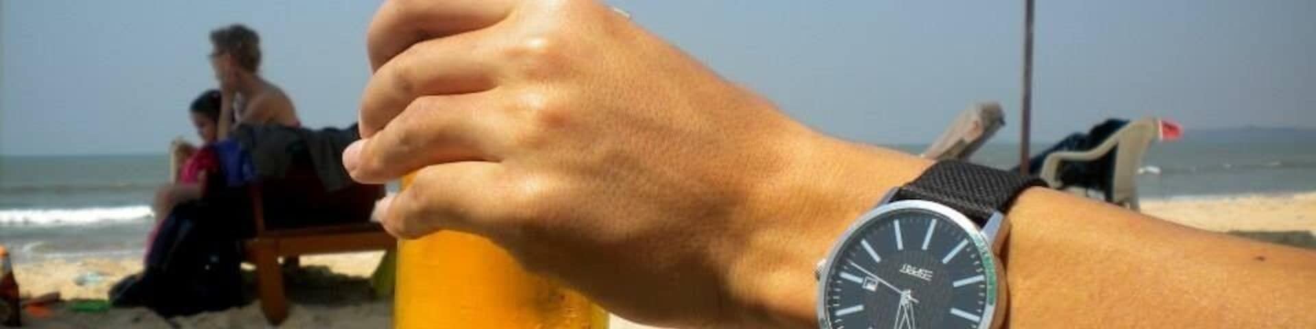 Uma pessoa segurando um copo de cerveja em uma praia, com um relógio no pulso e um guarda-sol verde ao fundo.