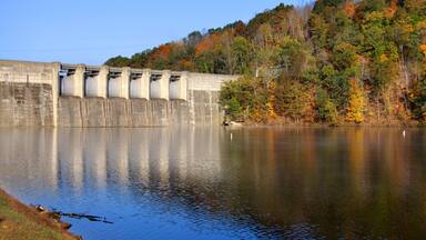 Uma barragem com várias verteduras e um lago calmo refletindo a folhagem outonal ao redor.