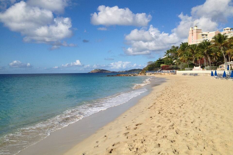 Uma praia arenosa com pegadas, céu azul límpido e uma ilha distante.