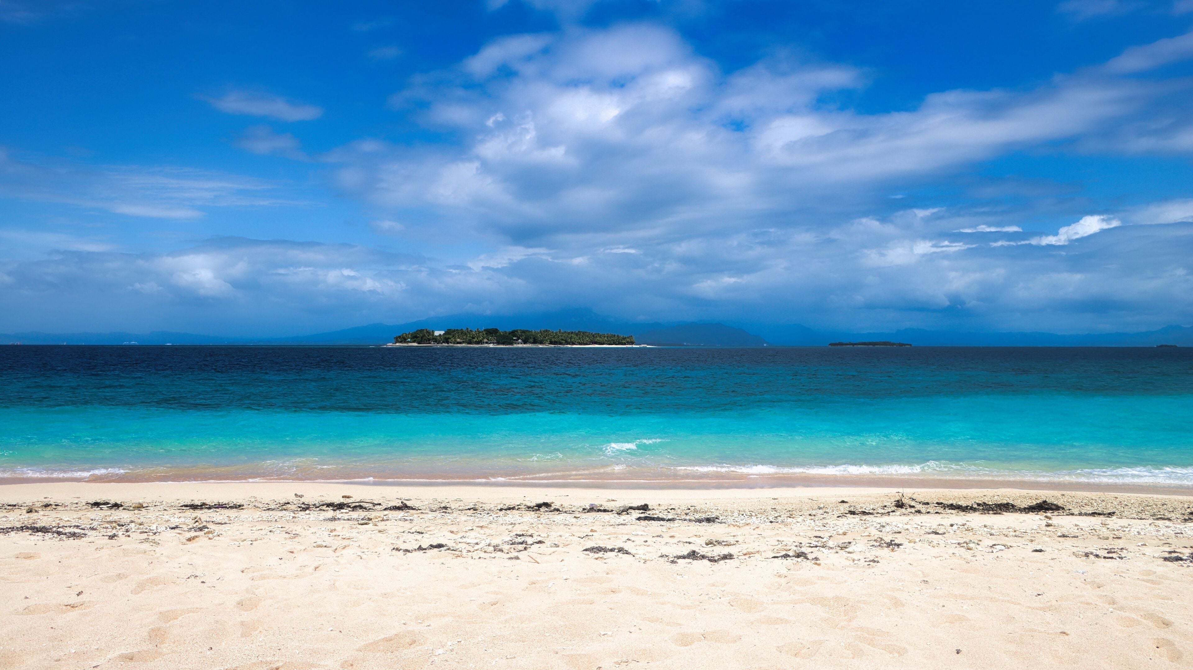 Uma praia com águas turquesa cristalinas, areal arenoso e uma pequena ilha com vegetação ao longe.