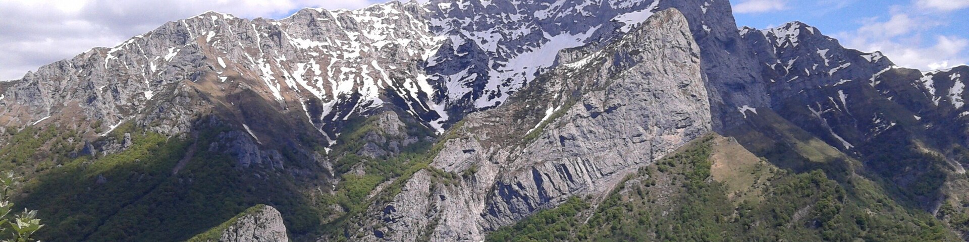 Uma cordilheira com picos nevados, vales densamente arborizados e um céu azul claro com nuvens esparsas.