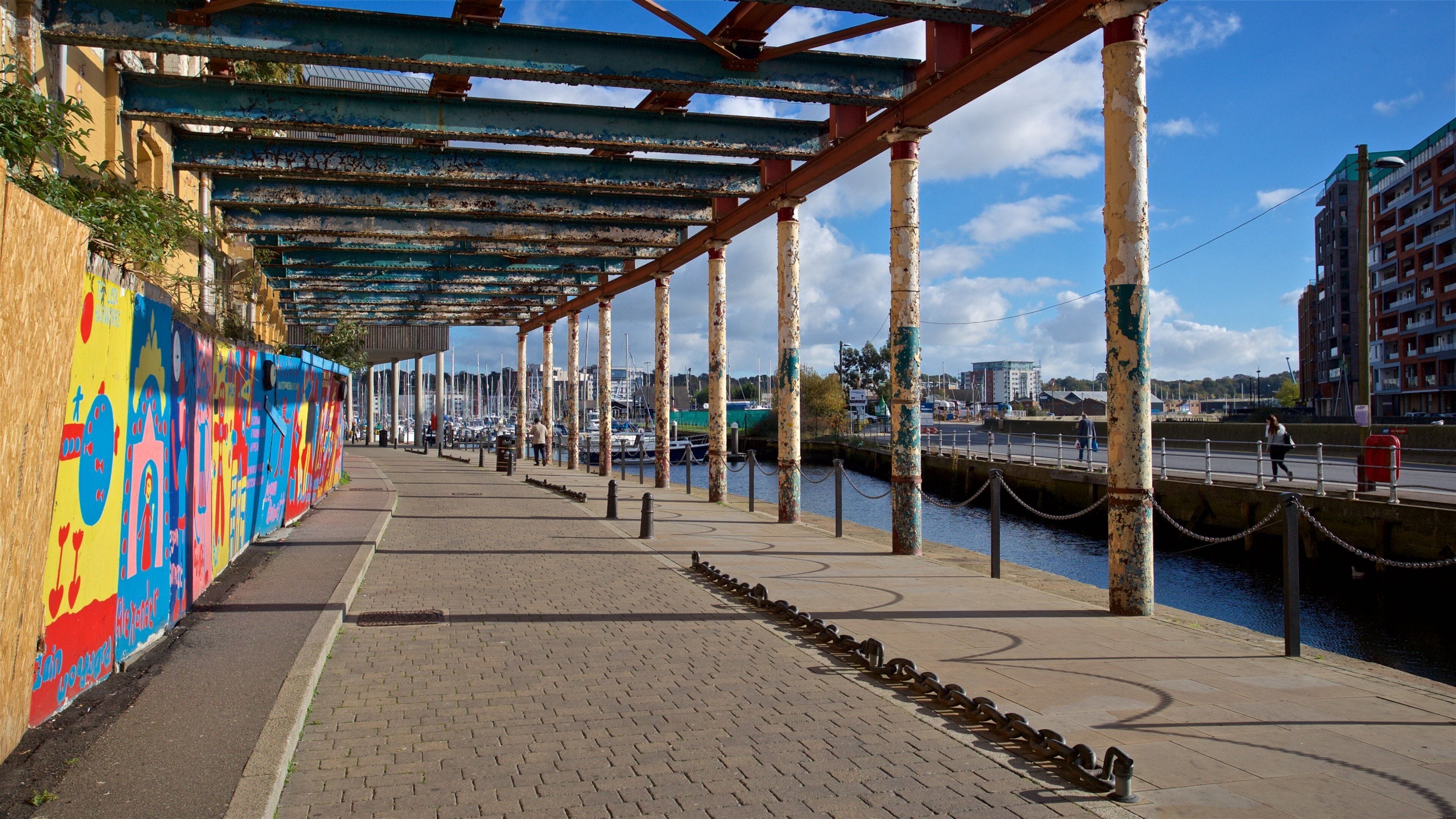 Um calçadão à beira-mar com um mural colorido, uma galeria coberta e um porto de embarcações ao fundo.