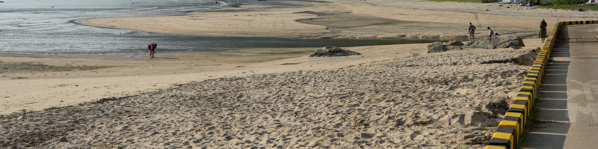 Uma praia de areia com uma barreira de segurança amarela e preta, uma montanha ao fundo e algumas pessoas à distância.