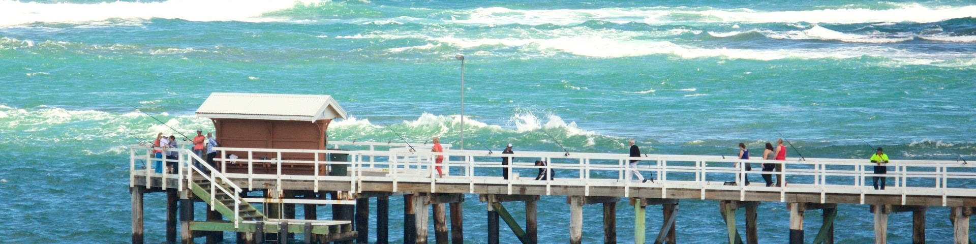 Um cais se estende para o oceano, com pessoas pescando e um pequeno edifício no final.