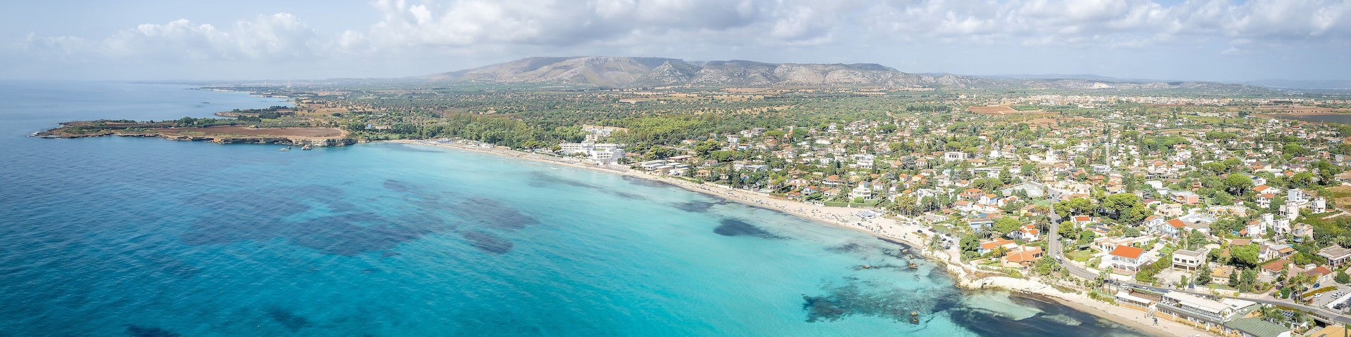 Uma vila costeira com praia, águas azul-claras e montanhas ao fundo.